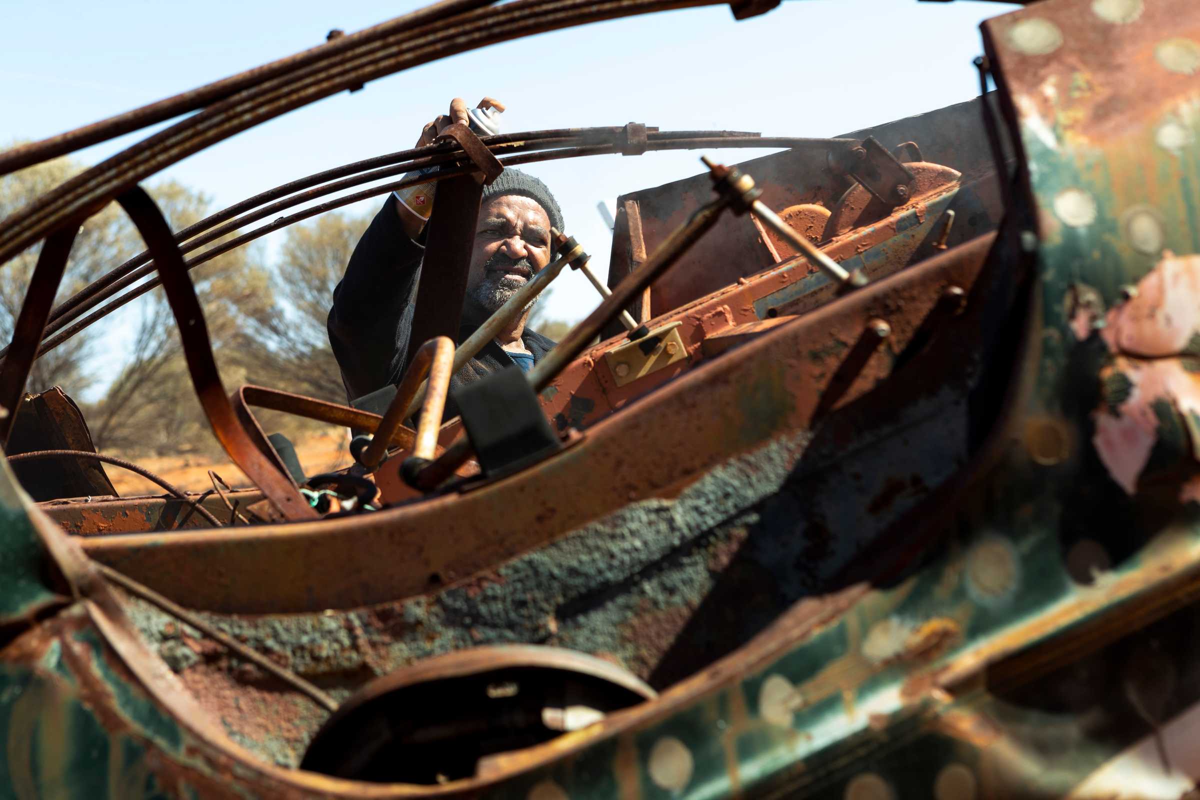 Image of the artist behind the wreckage of a car with a can of spray paint